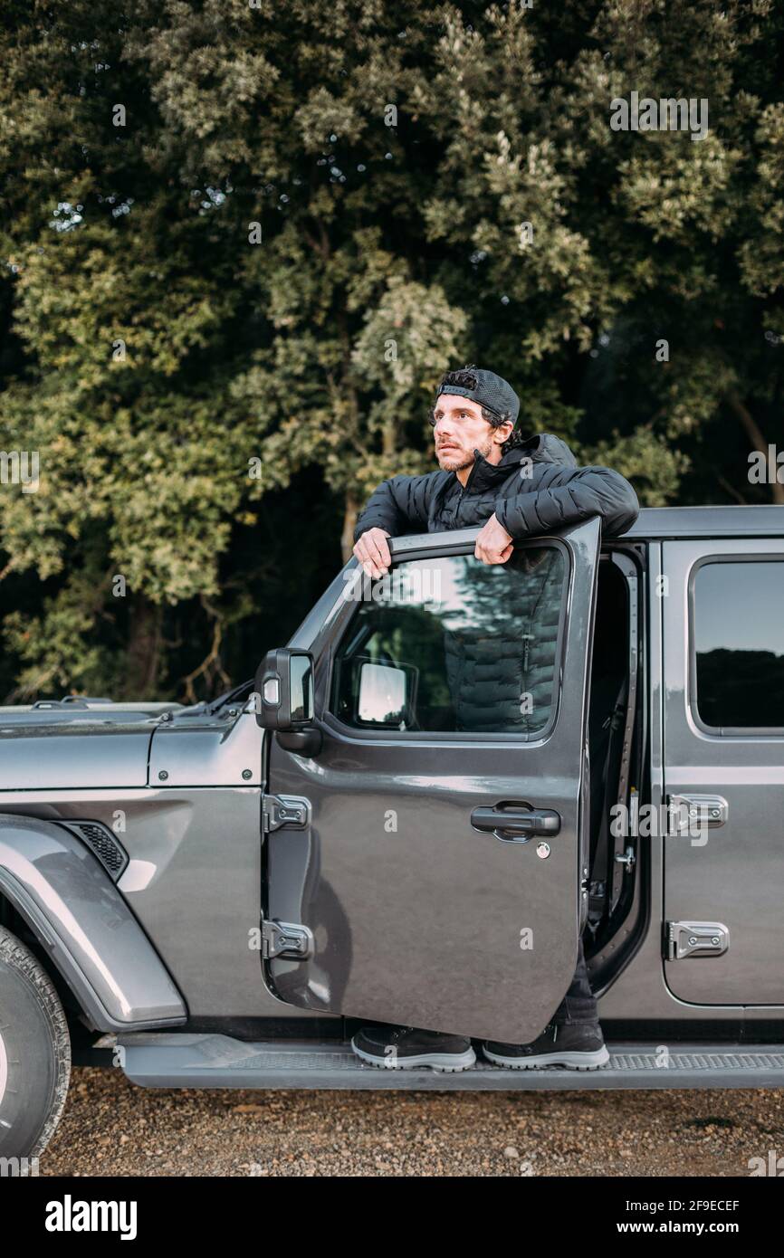 Side view of an adventurer leaning on the door of an off-road car while ...