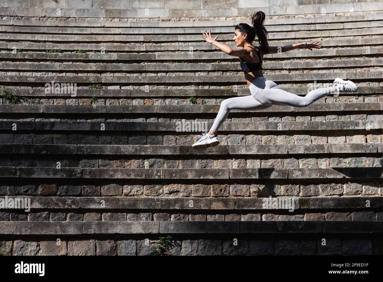 Side view of fit female in sportswear jumping in splits above stone ...