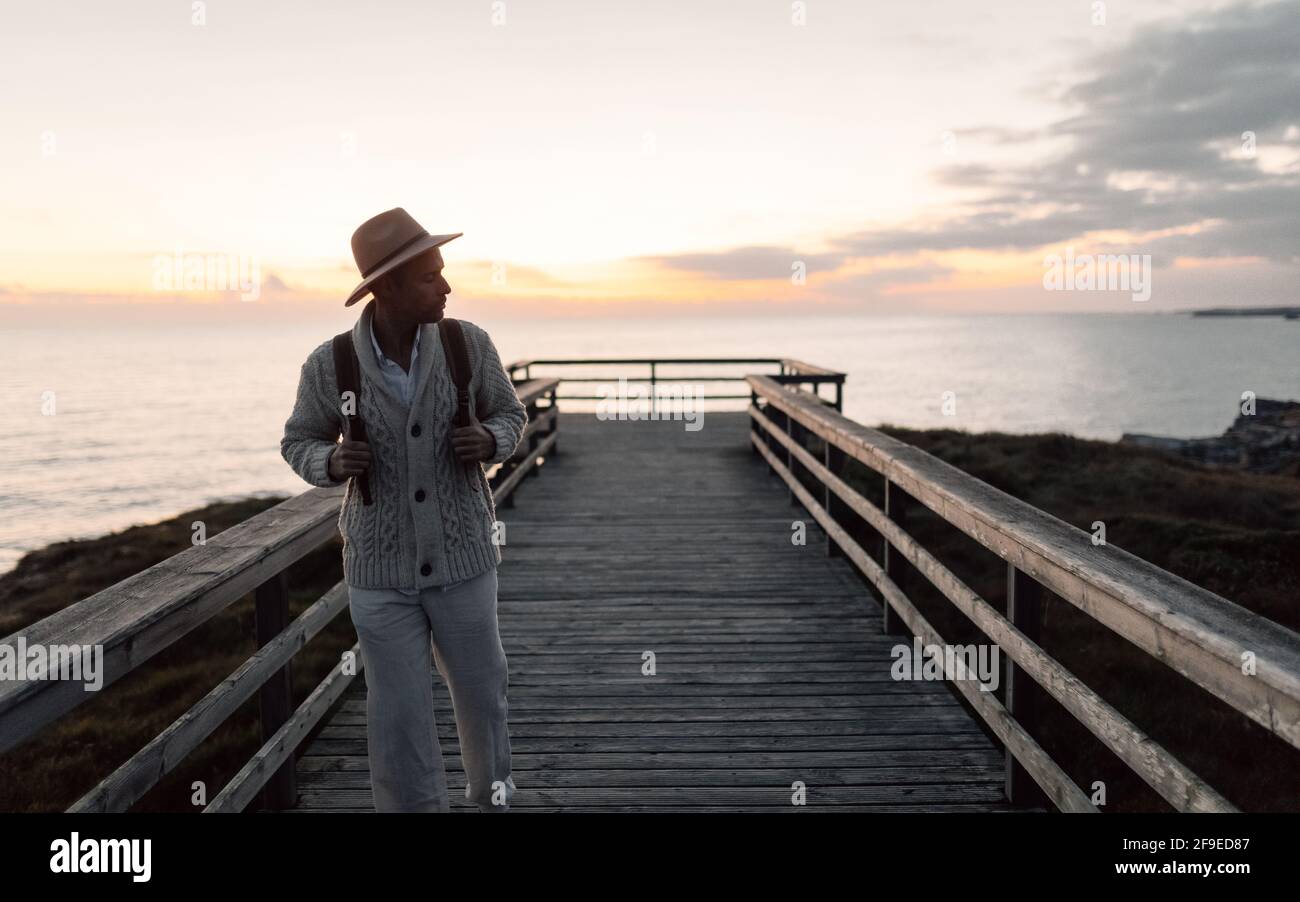 man with backpack and hat standing on a walkway looking at the sea ...