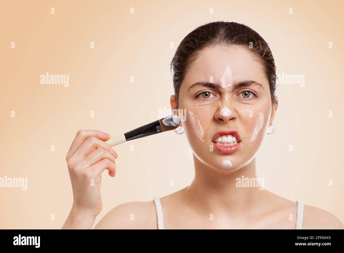 Portrait of a young angry woman applying a skin care mask with a brush ...