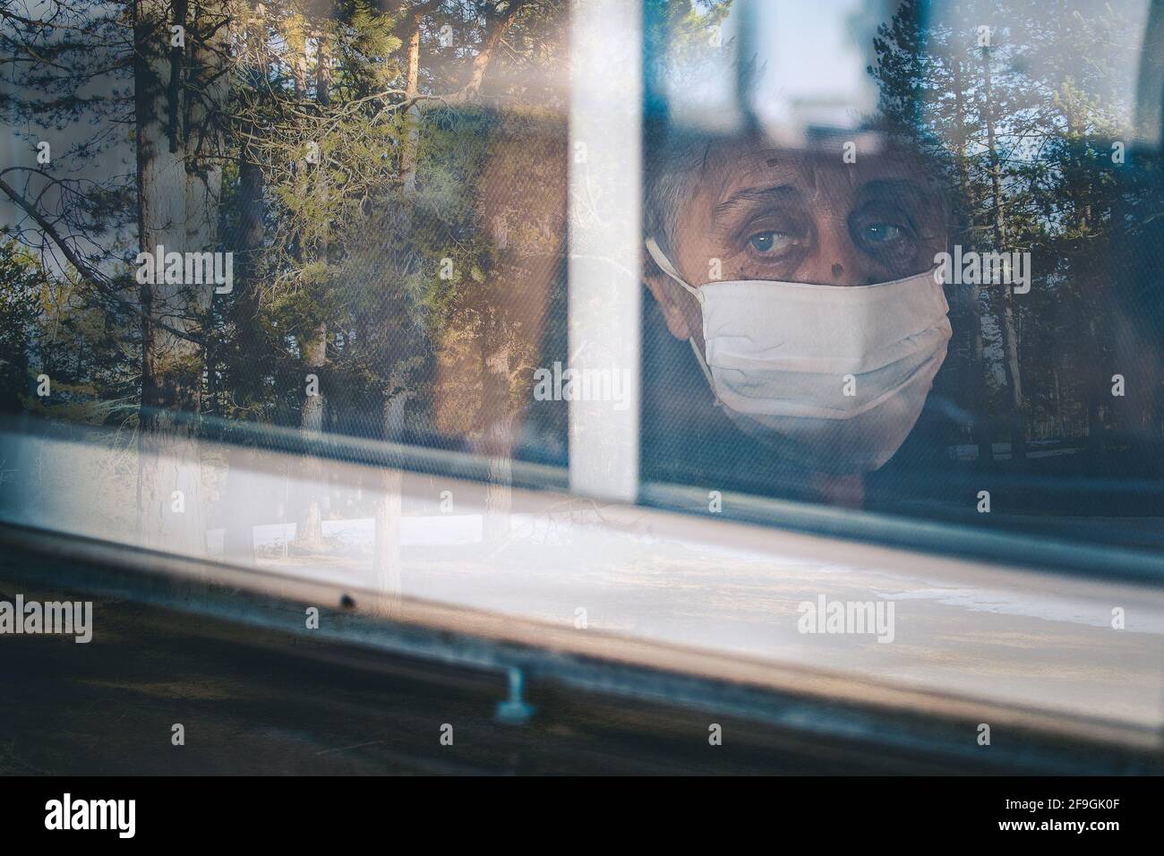 Elderly woman, grandma wearing a mask looking through the window. View ...