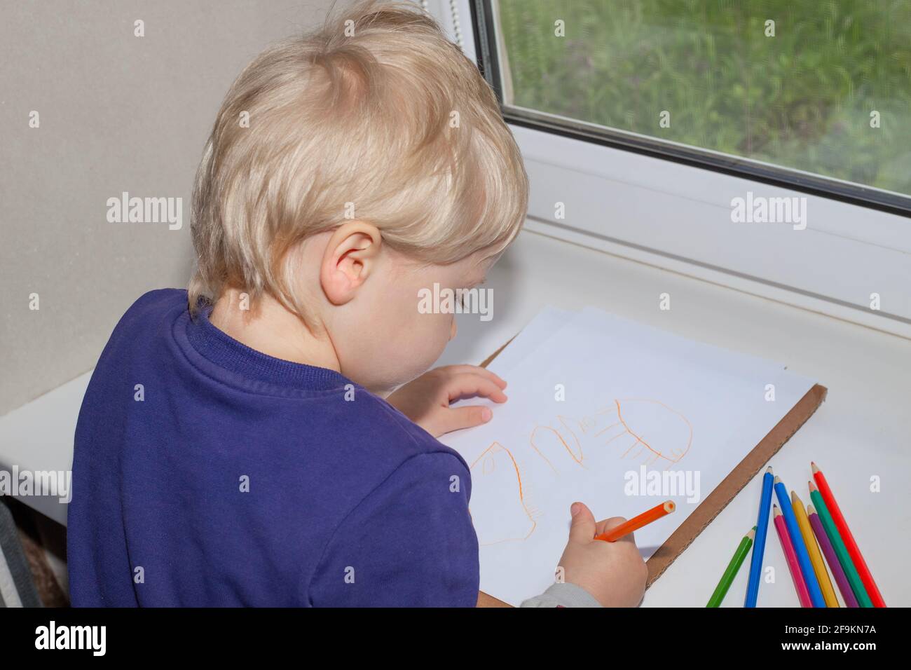 Boy blond draws with a pencil on a white sheet of paper while sitting ...