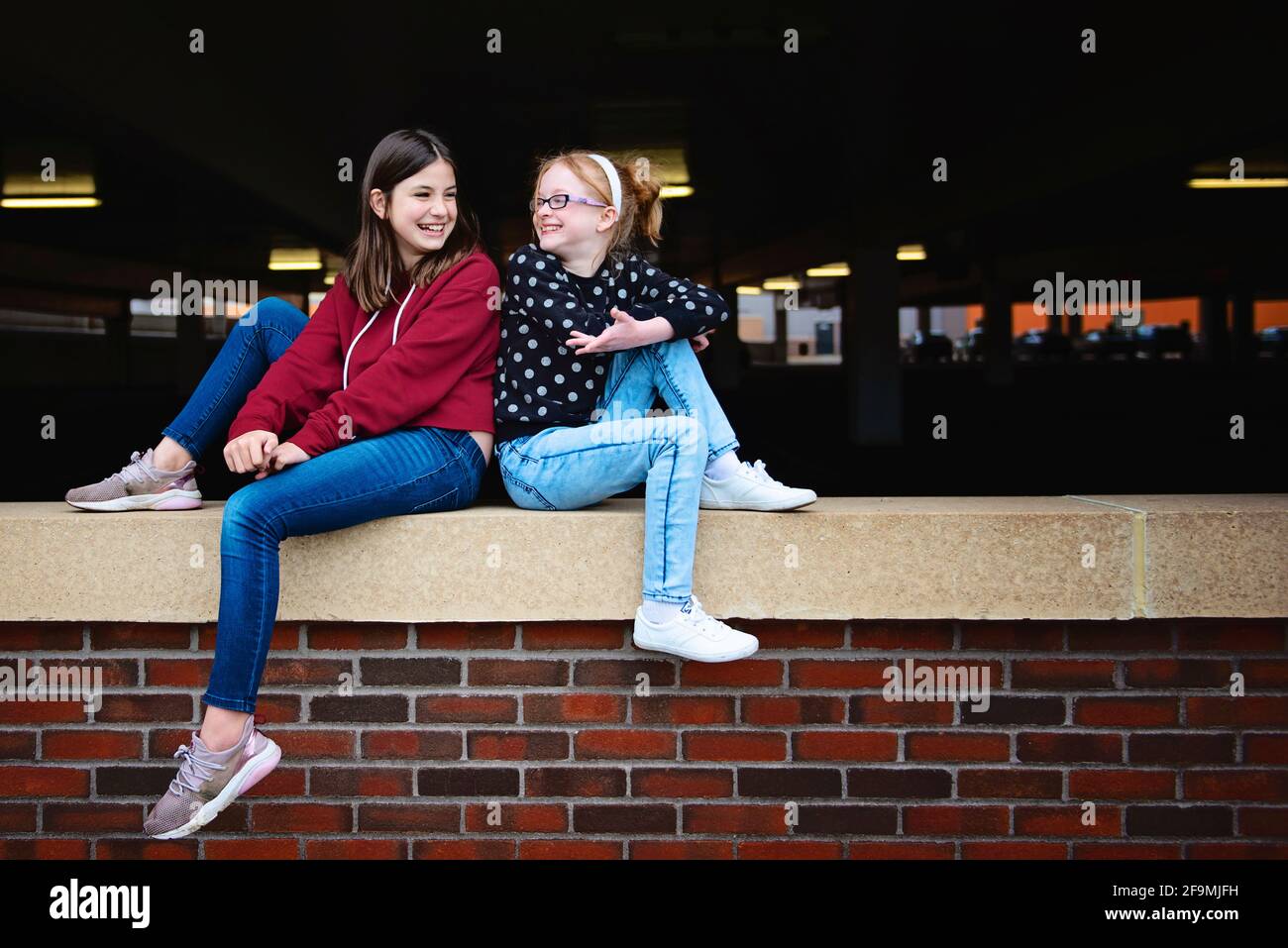 Two happy tween girls sitting on a brick wall together talking Stock ...