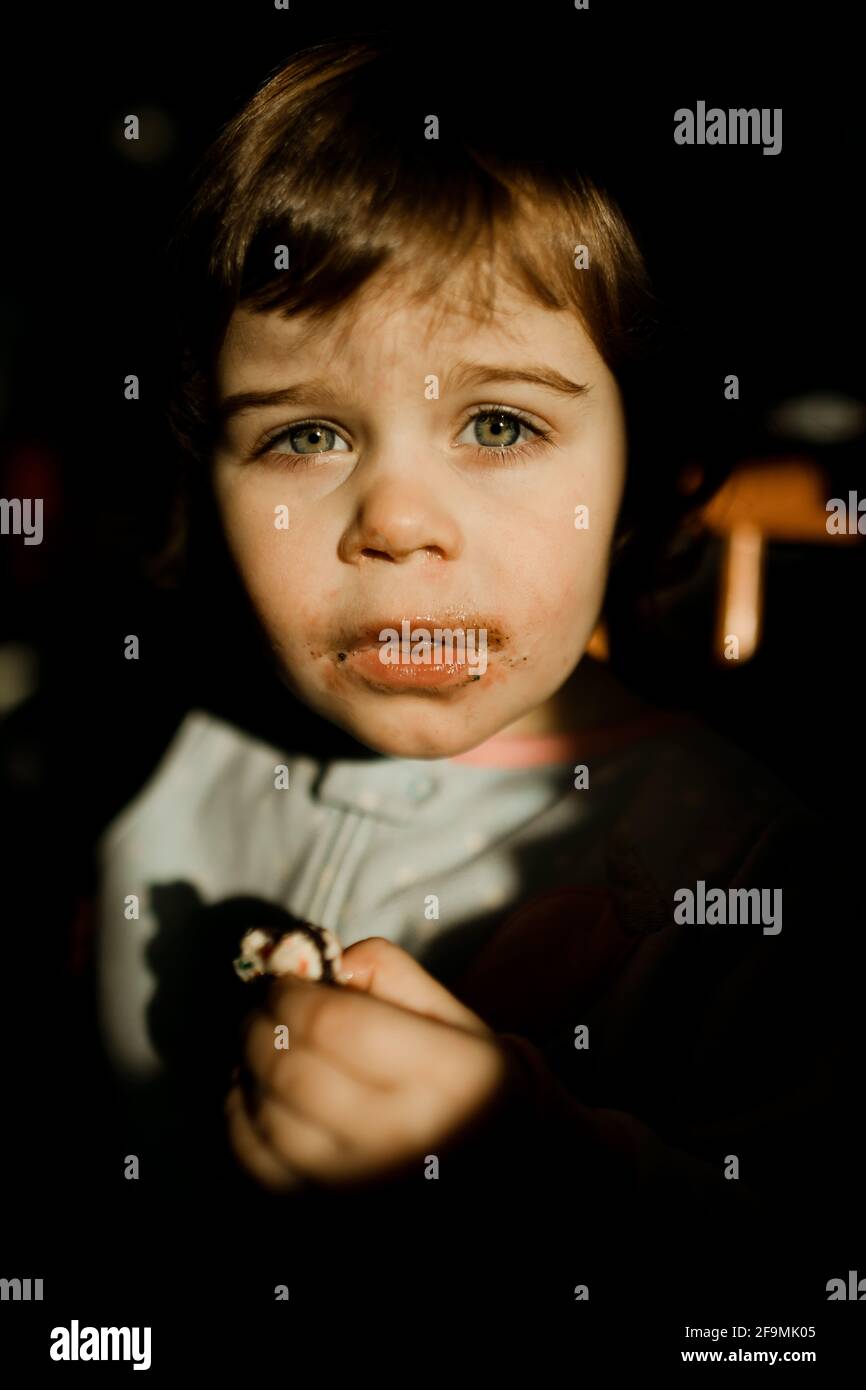 Young girl eating a cookie on kitchen chair with crumbs on her face ...