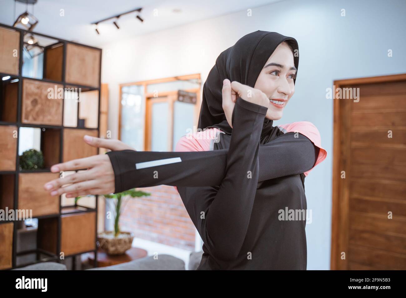 A Muslim woman in sports clothes stands stretching with one hand ...
