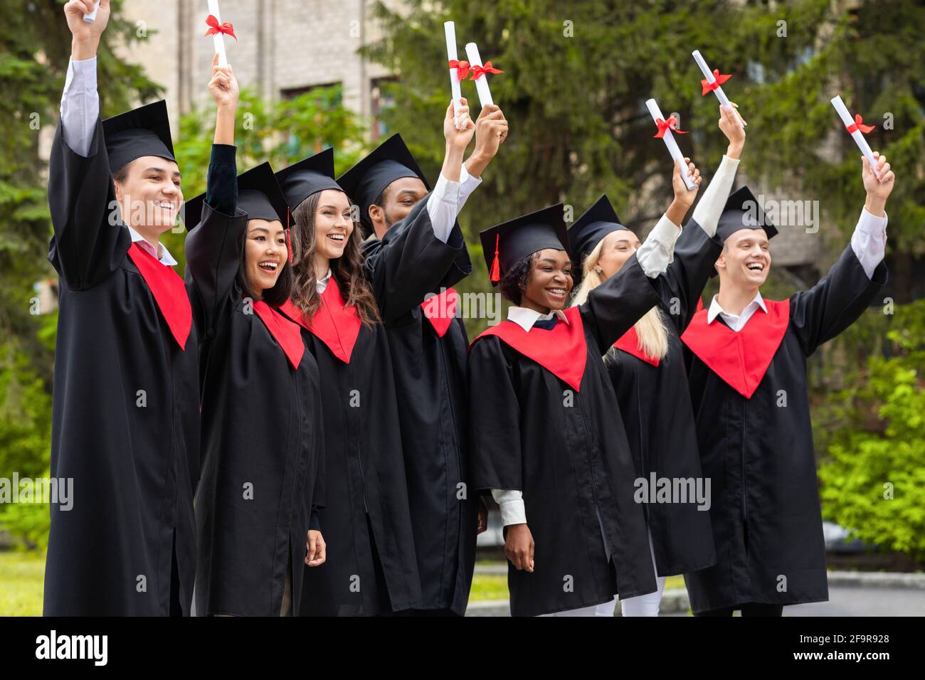Emotional multiethnic students in graduation costumes raising diplomas ...