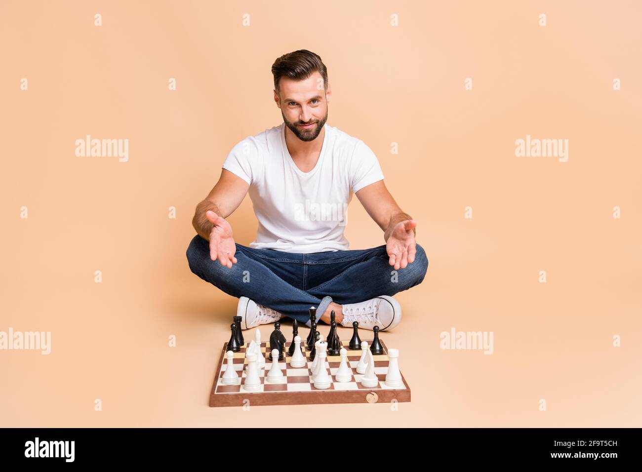 Portrait of nice cheery guy sitting on floor playing chess inviting you ...