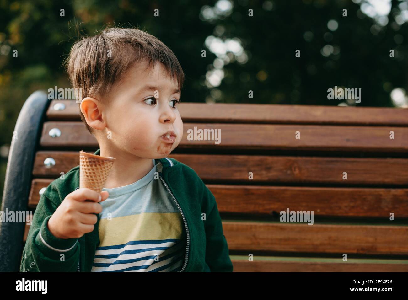 Caucasian boy is eating ice cream sitting on a bench in park looking at ...