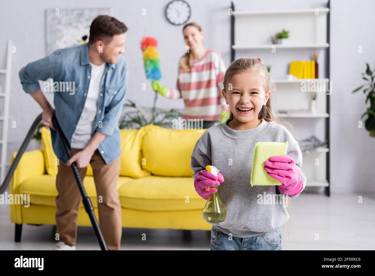 Smiling kid in rubber gloves holding detergent and rag near parents on ...