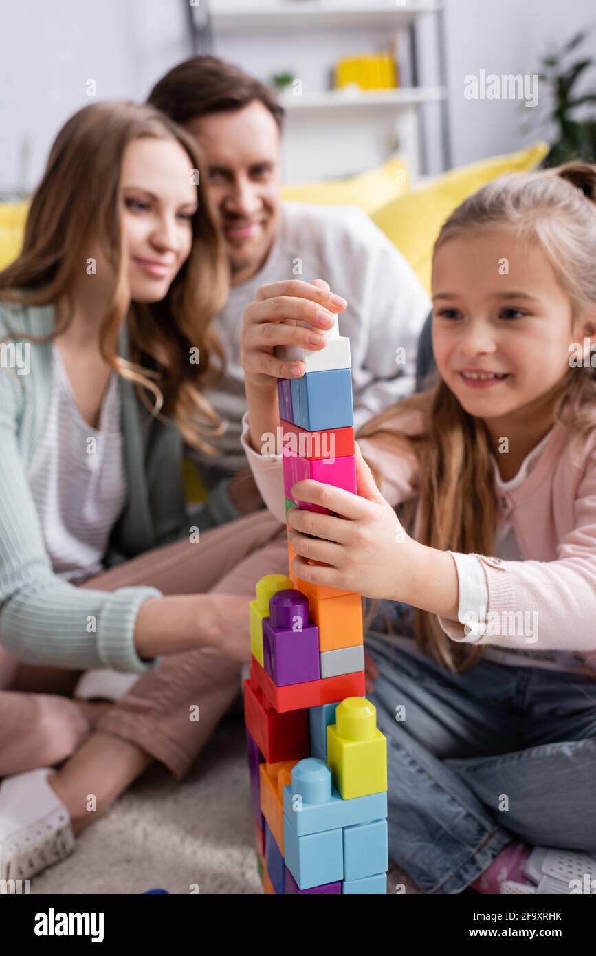 Smiling girl on blurred background playing building blocks near parents ...