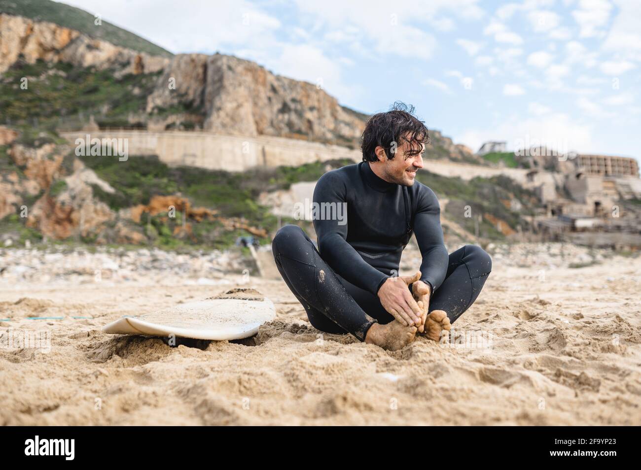 Young surfer man wearing diving suit sitting on the sand next to his ...