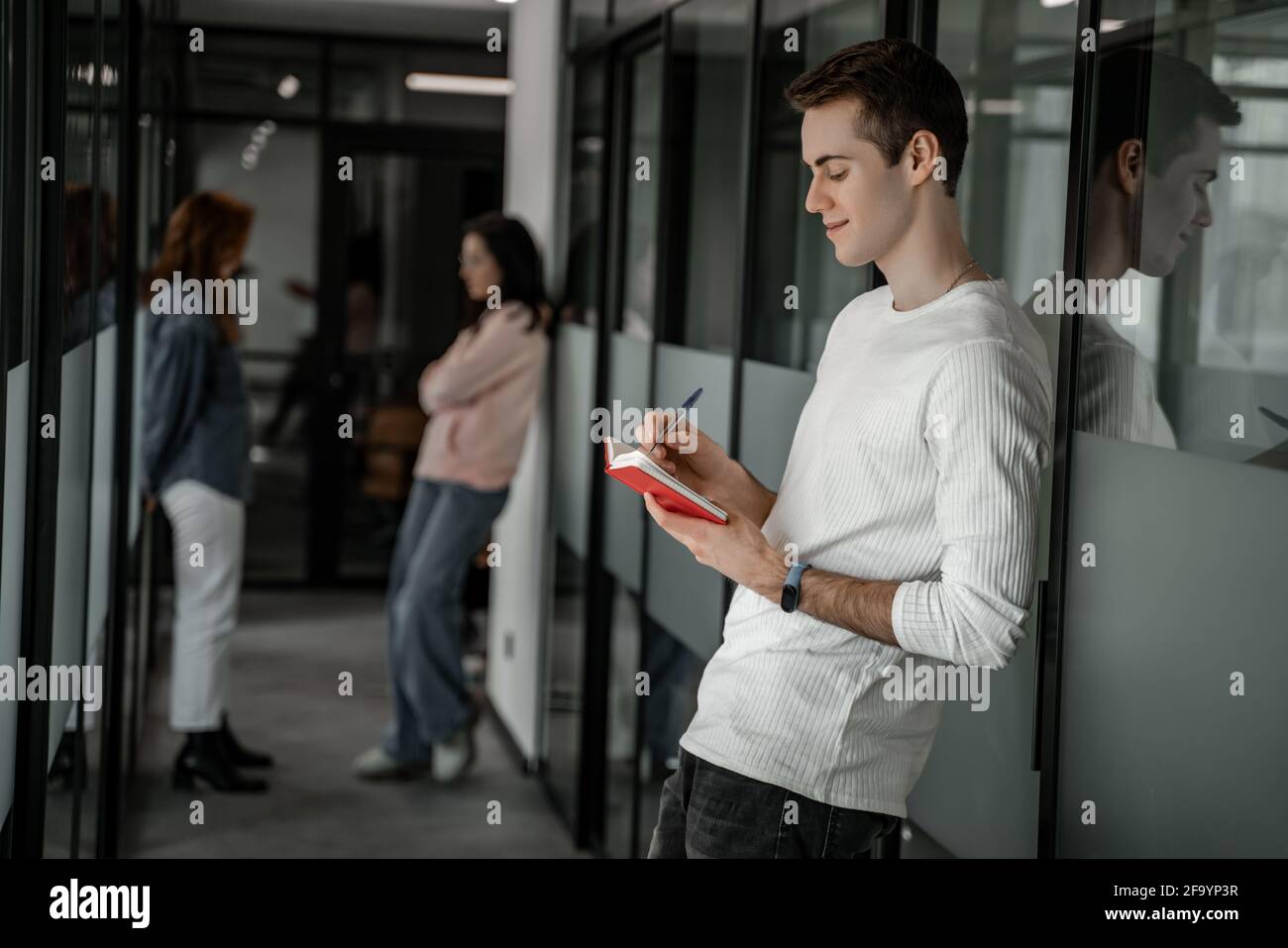smiling young student standing in hall and writing in notebook near ...