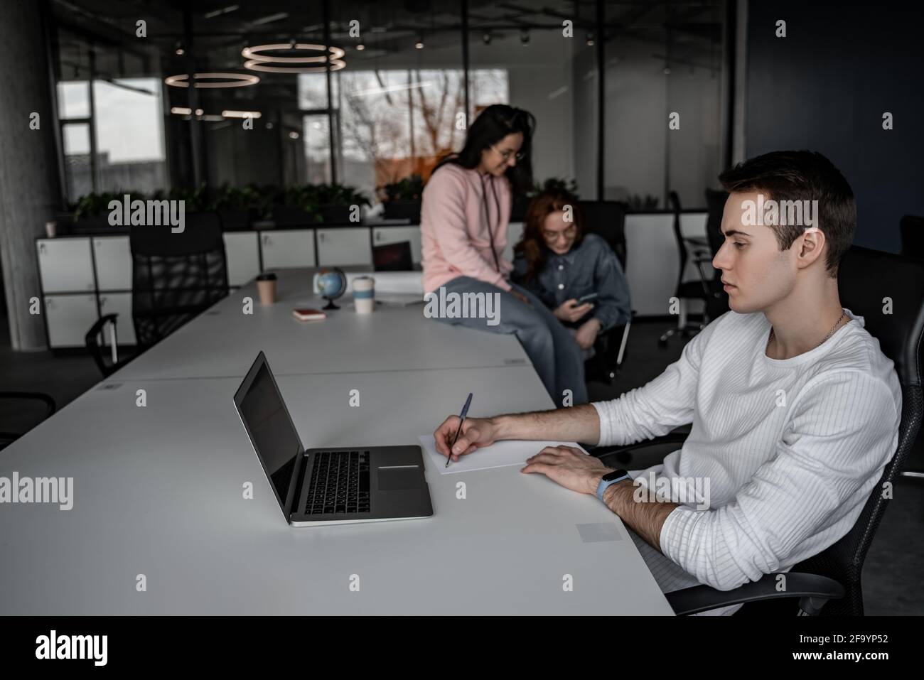 young student writing on paper while looking at laptop near women on ...