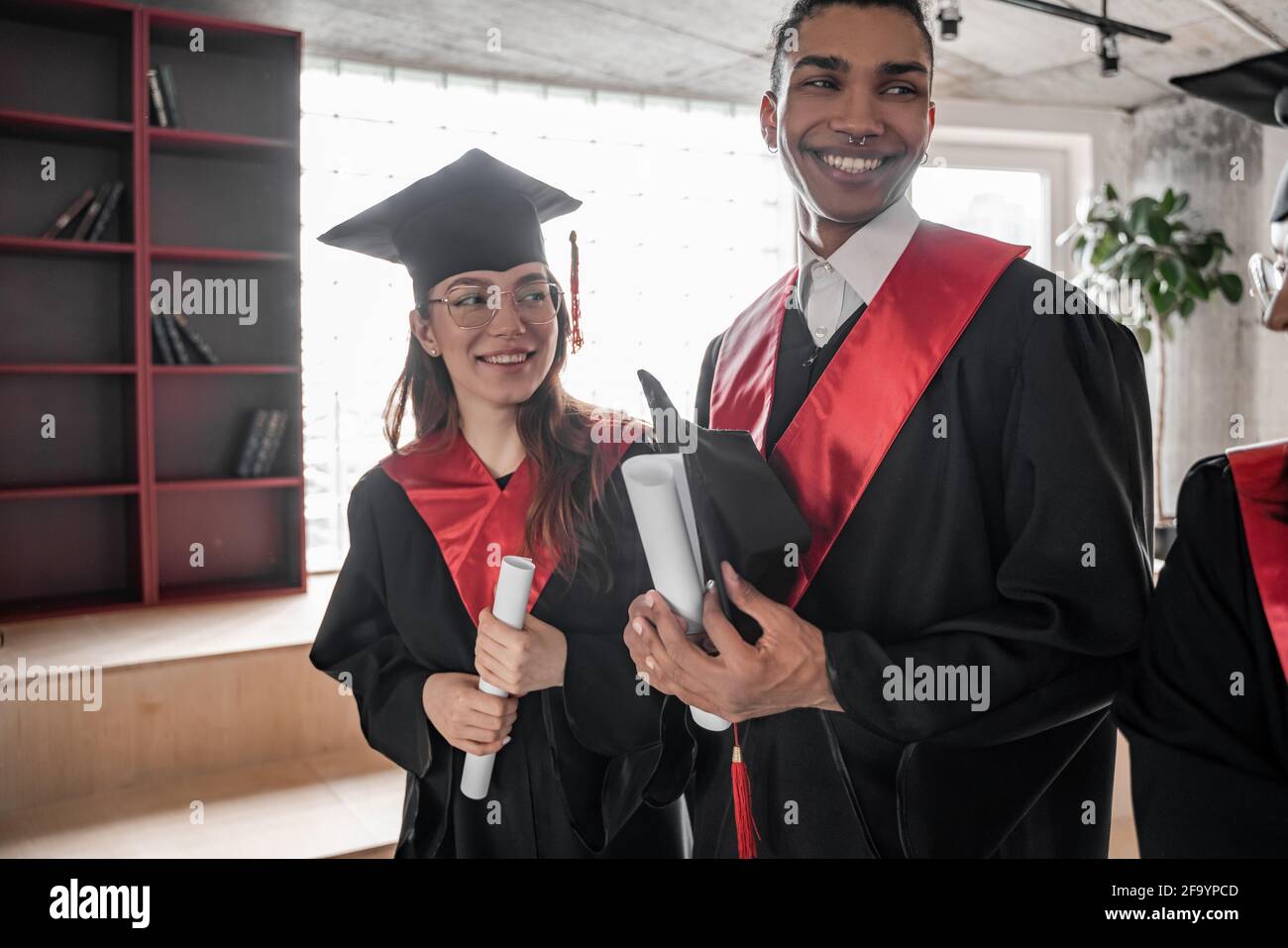 cheerful interracial students in graduation gowns and caps holding ...