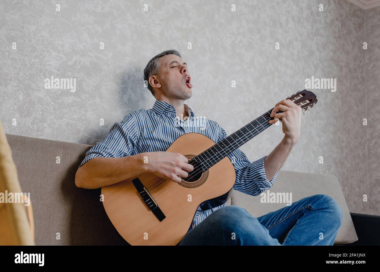 A young man plays the guitar sitting on a sofa in a bright living room ...
