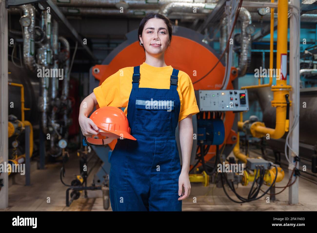 A young smiling female worker in uniform holds a safety helmet in her ...