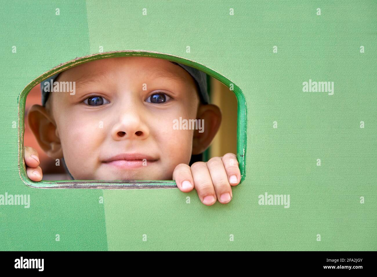 Cute happy little boy looks through small window in green wooden house ...