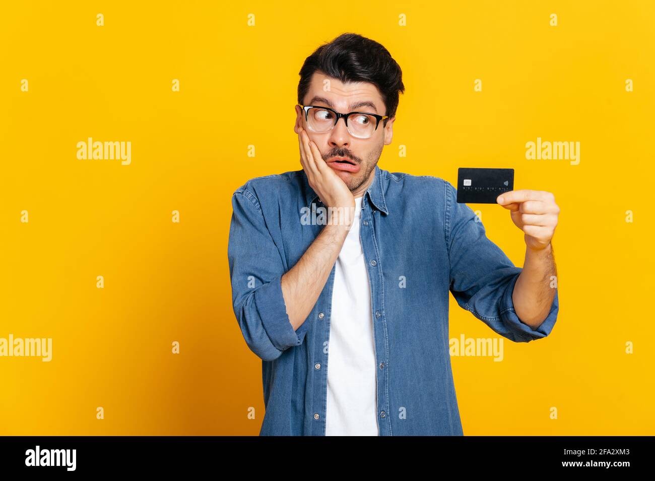 Surprised puzzled caucasian guy with glasses holding a credit card in ...
