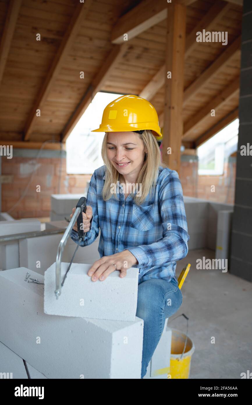 pretty young female bricklayer with yellow safety helmet is sawing ...
