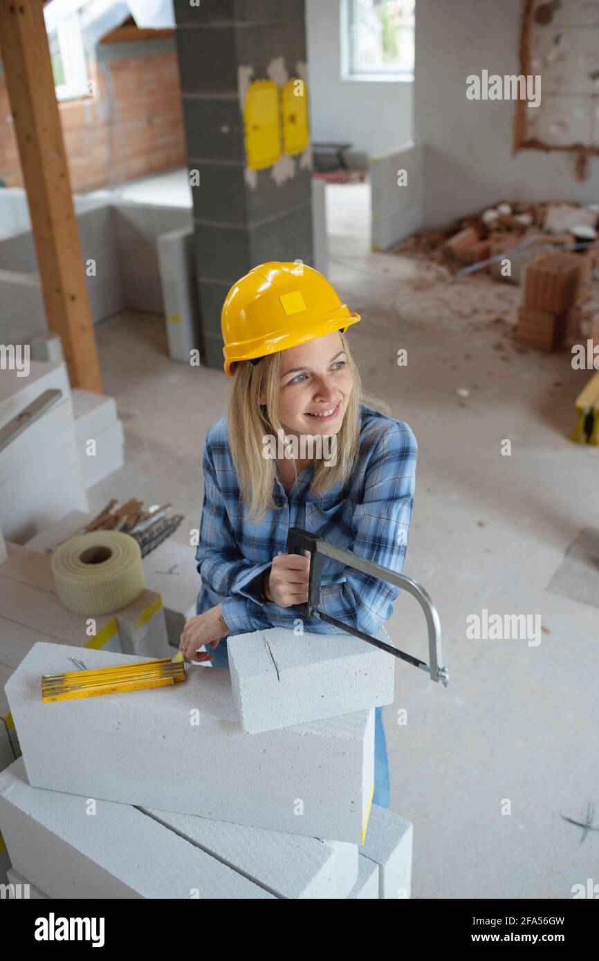 pretty young female bricklayer with yellow safety helmet is sawing ...