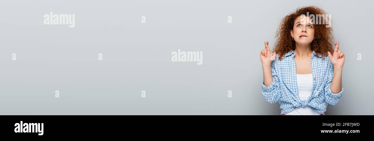 tense woman with crossed fingers looking up on grey background, banner ...