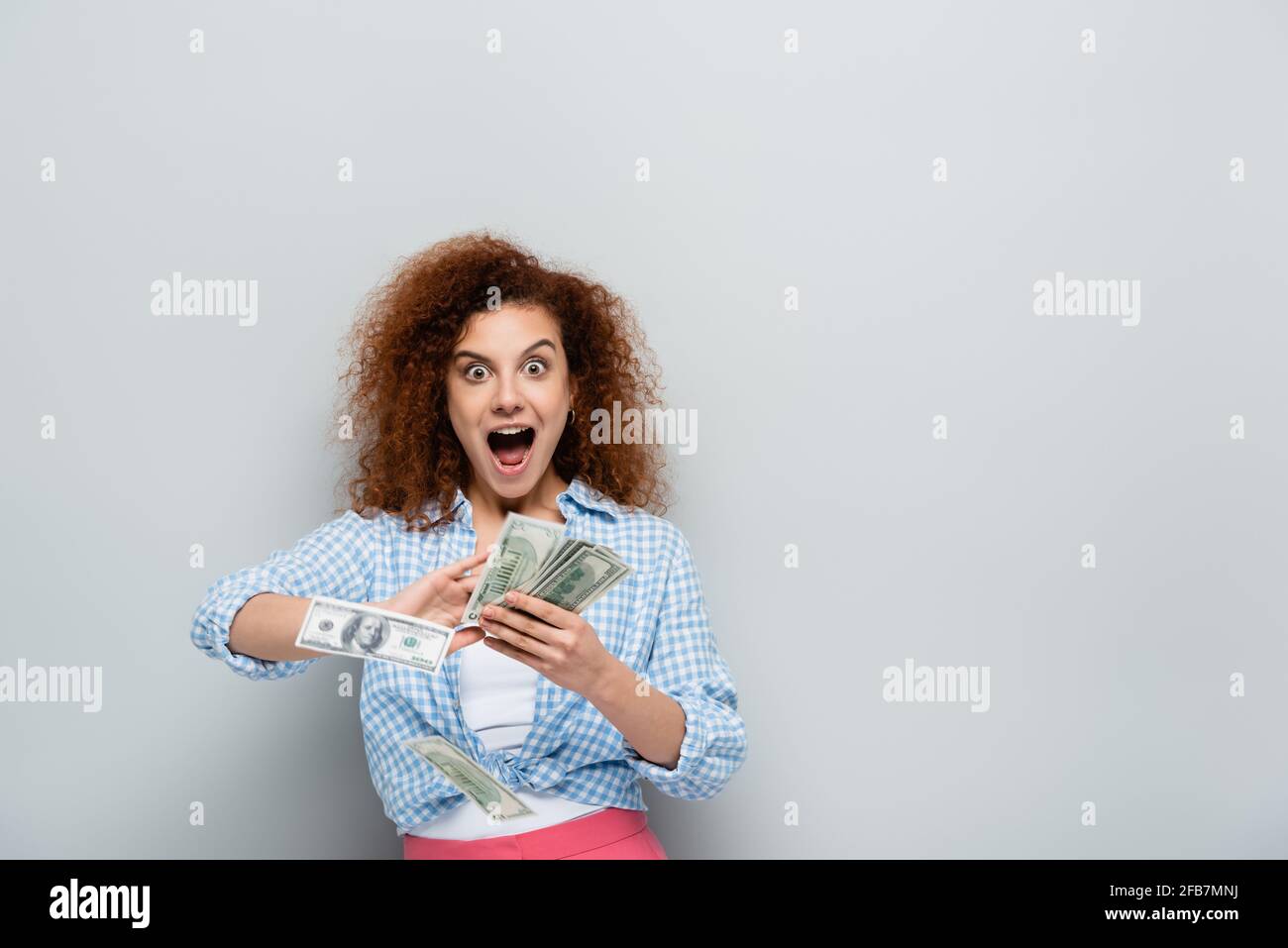 astonished woman screaming while counting dollar banknotes on grey ...
