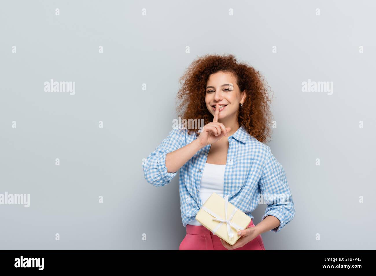 cheerful woman showing shh gesture while holding gift box on grey ...