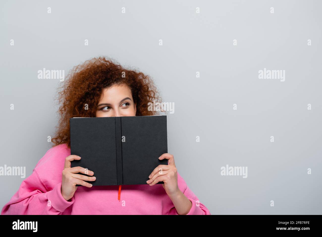 young woman with curly hair obscuring face with notebook isolated on ...