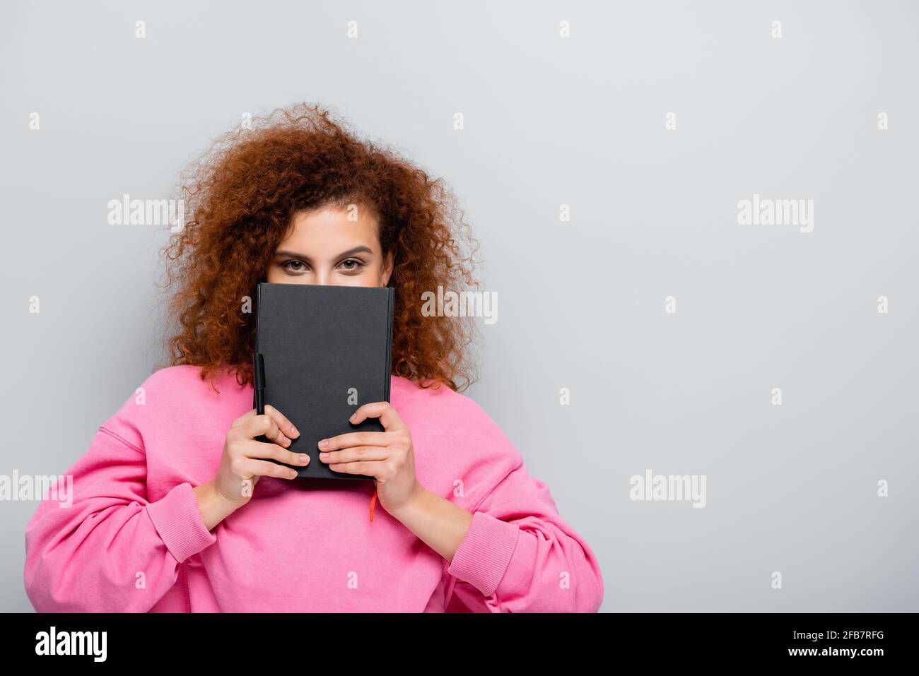 curly woman obscuring face with notebook while looking at camera ...
