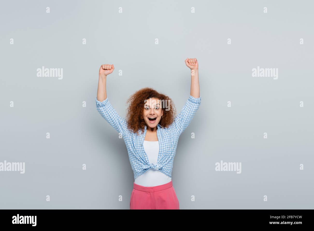 astonished woman with curly hair showing rejoice gesture on grey ...