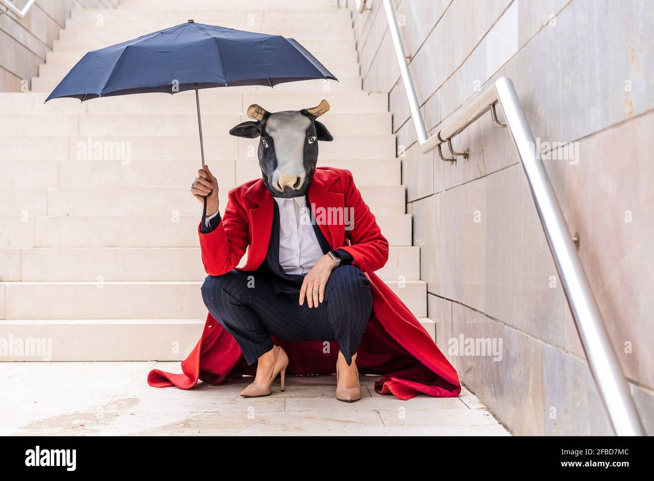 Businesswoman wearing bull mask holding umbrella while crouching near ...