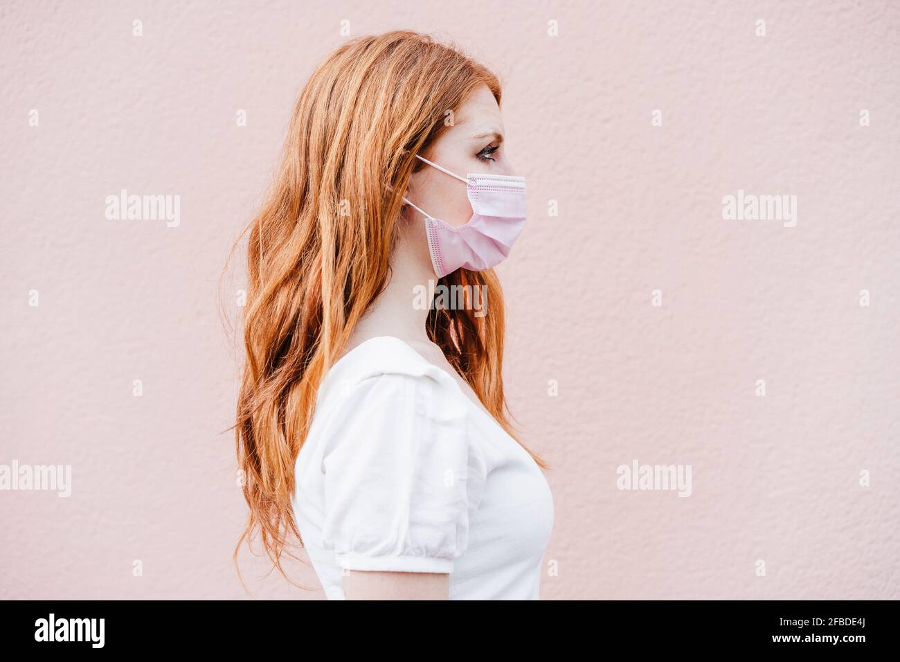 Young redhead woman wearing mask by pink wall during COVID-19 Stock ...