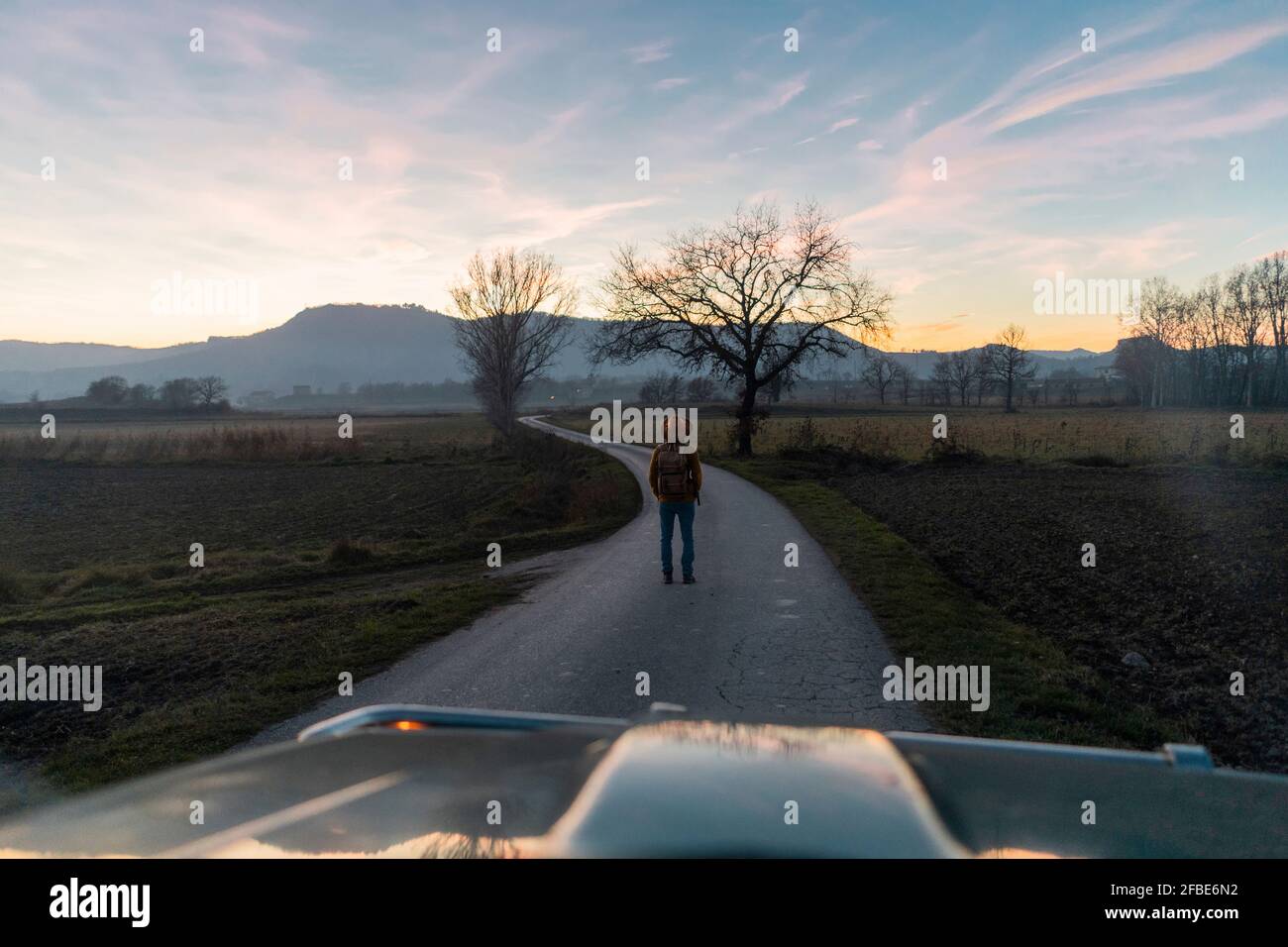 Man standing in front of car on country road during sunset Stock Photo ...