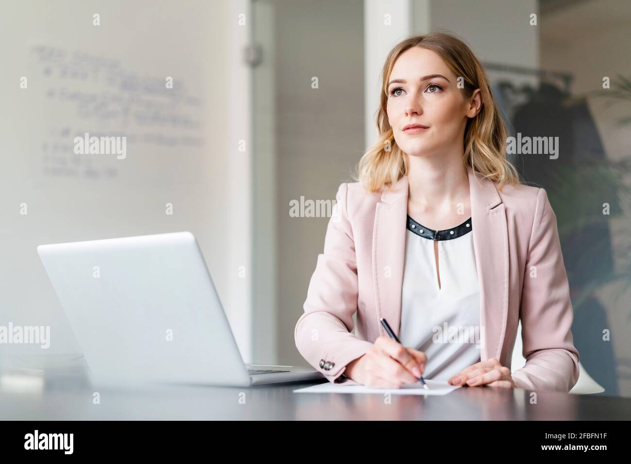 Businesswoman contemplating while writing on document by laptop in ...