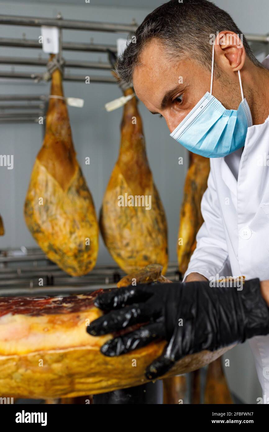 Male butcher in protective face mask cutting ham at shop Stock Photo ...
