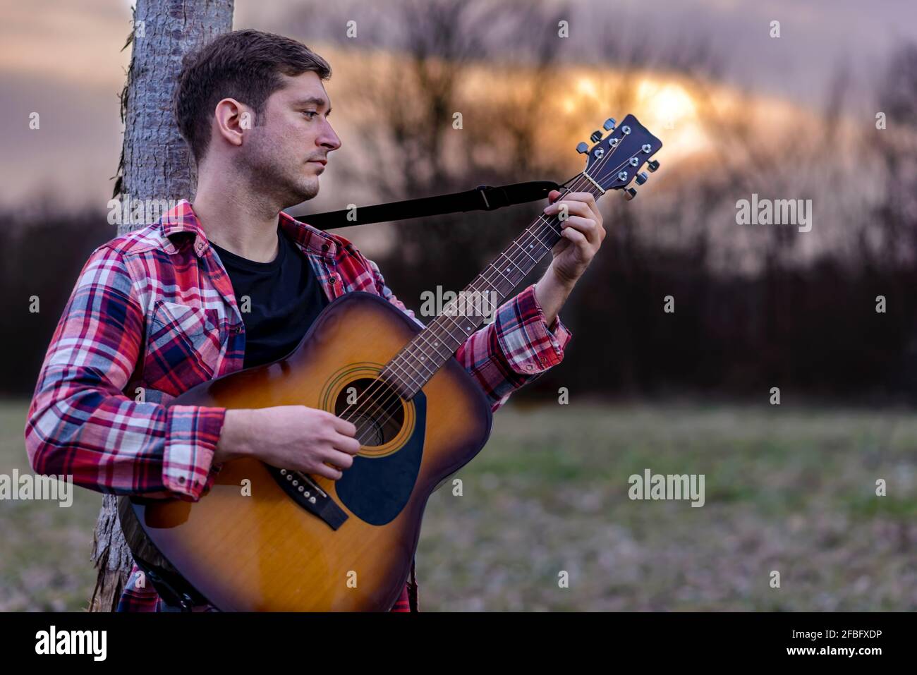 A handsome young man leaning against a tree plays an acoustic guitar in ...