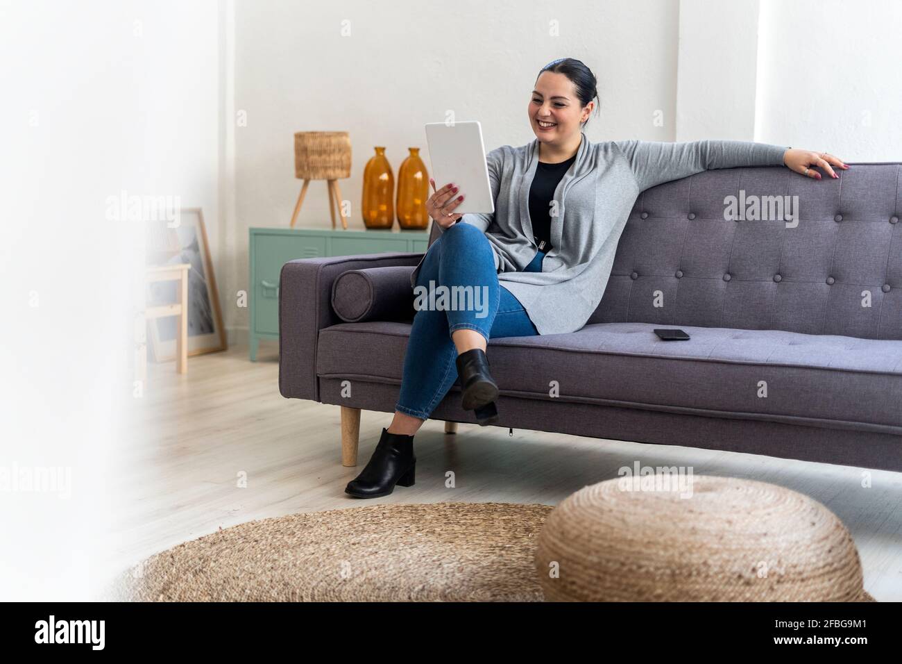 Smiling woman using digital tablet while sitting on sofa in living room ...