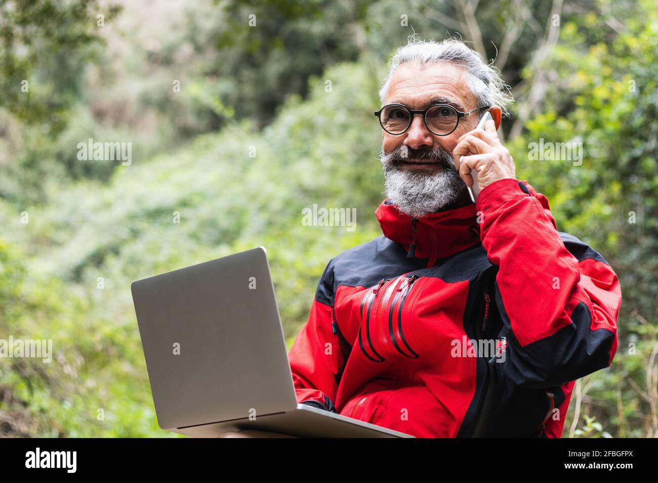 Smiling male explorer with laptop talking on mobile phone in forest ...