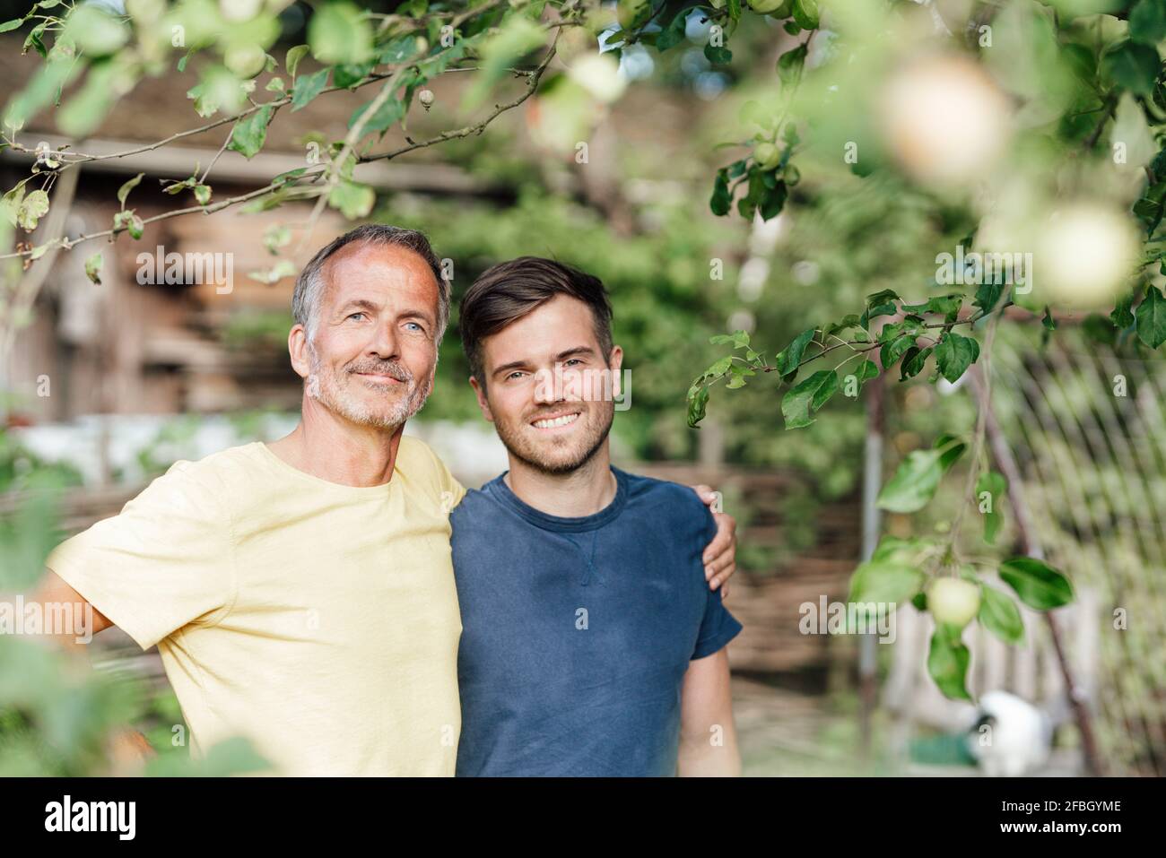 Confident father with arm around son standing under tree in backyard ...