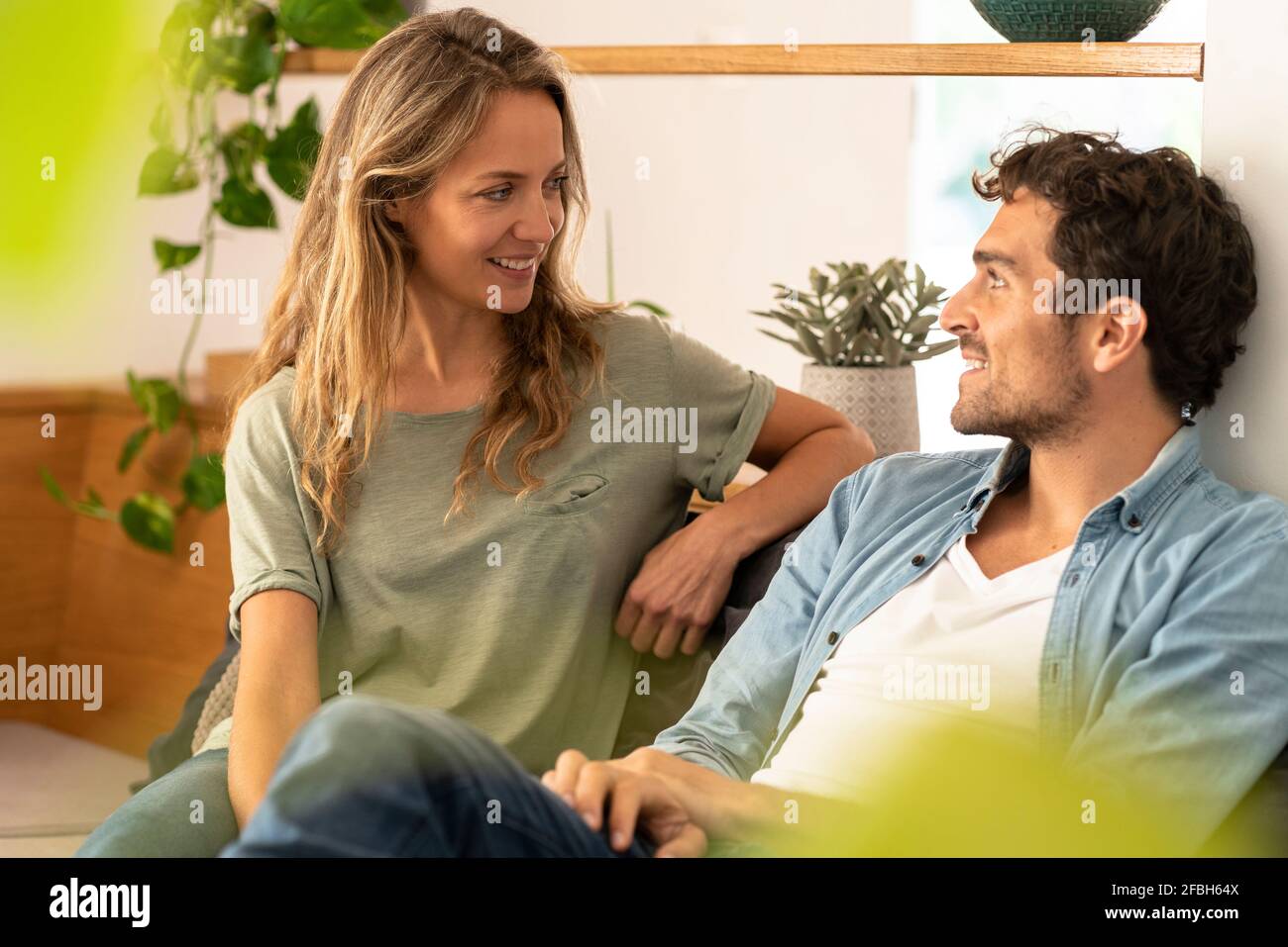 Smiling girlfriend looking at boyfriend while sitting on sofa in living ...