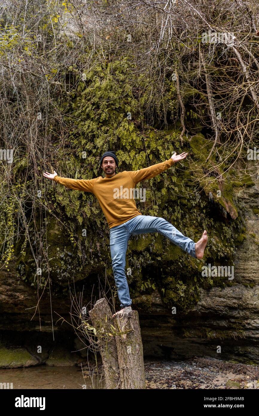 Carefree man with arms outstretched standing on one leg at tree stump ...