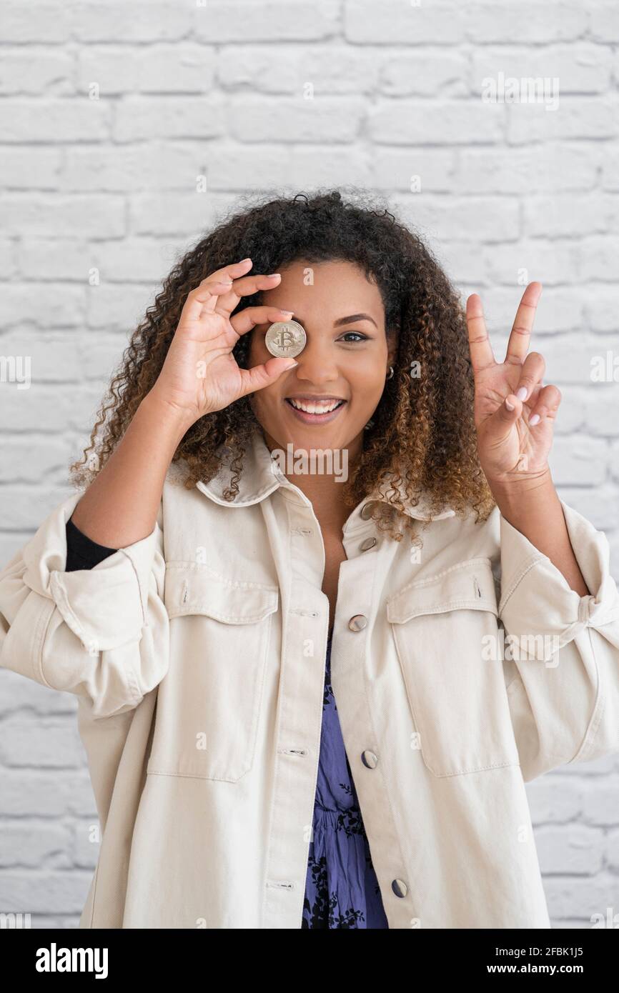 Smiling young woman showing peace sign while covering eye with bitcoin ...