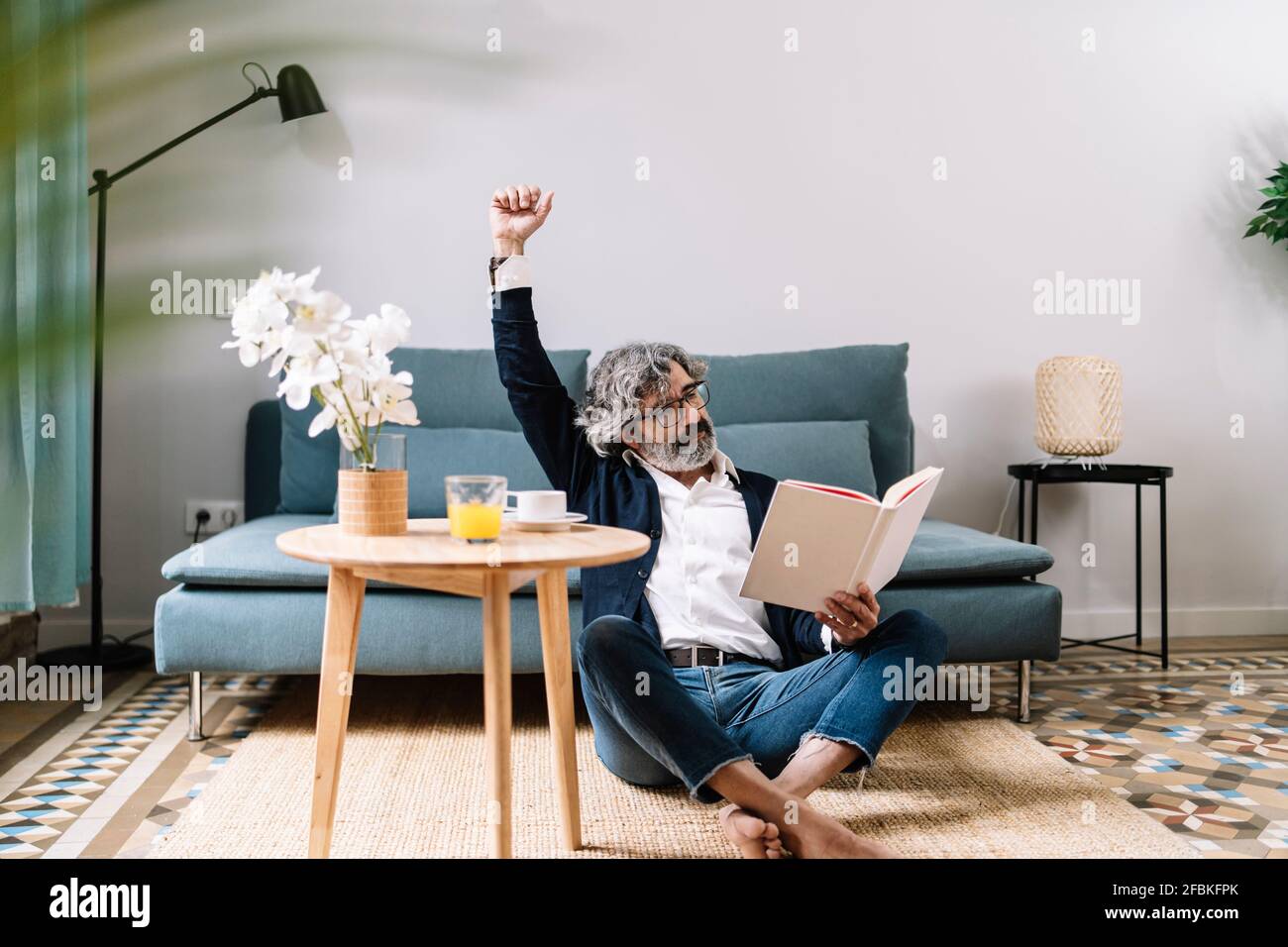 Tired senior man stretching arm while reading book by sofa at home ...