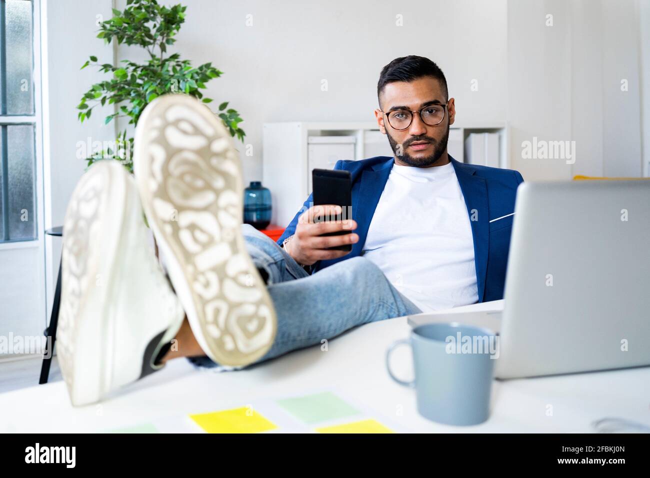 Businessman with smart phone relaxing with feet up on desk at office ...