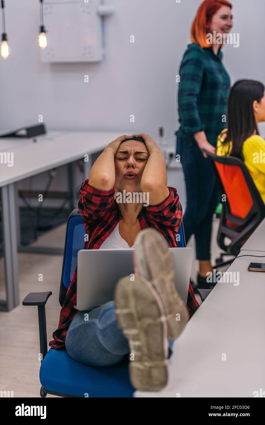 A nervous female office worker with her legs on her desk and her laptop ...