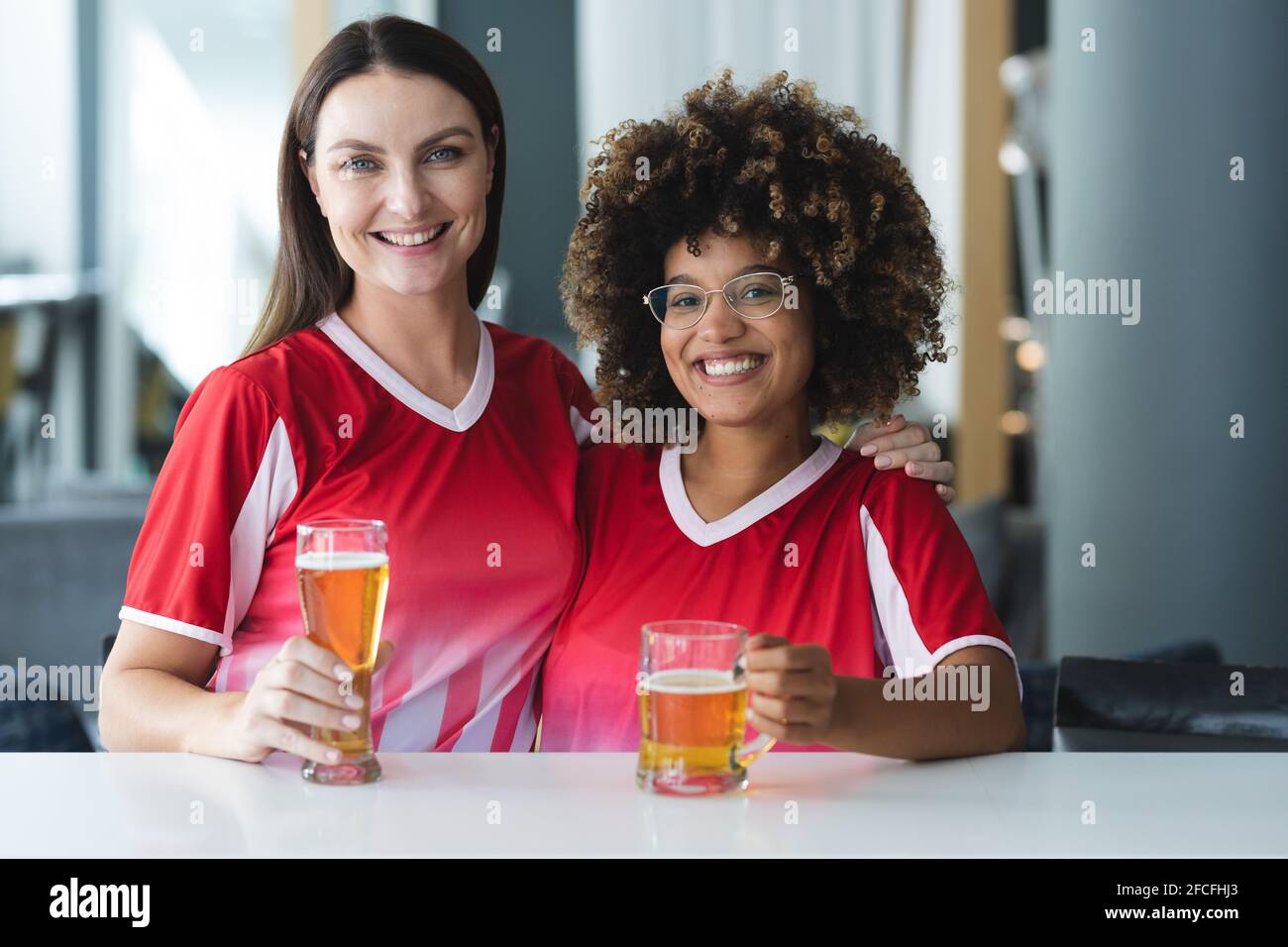 Portrait of two diverse female sports fans having beer smiling at bar ...