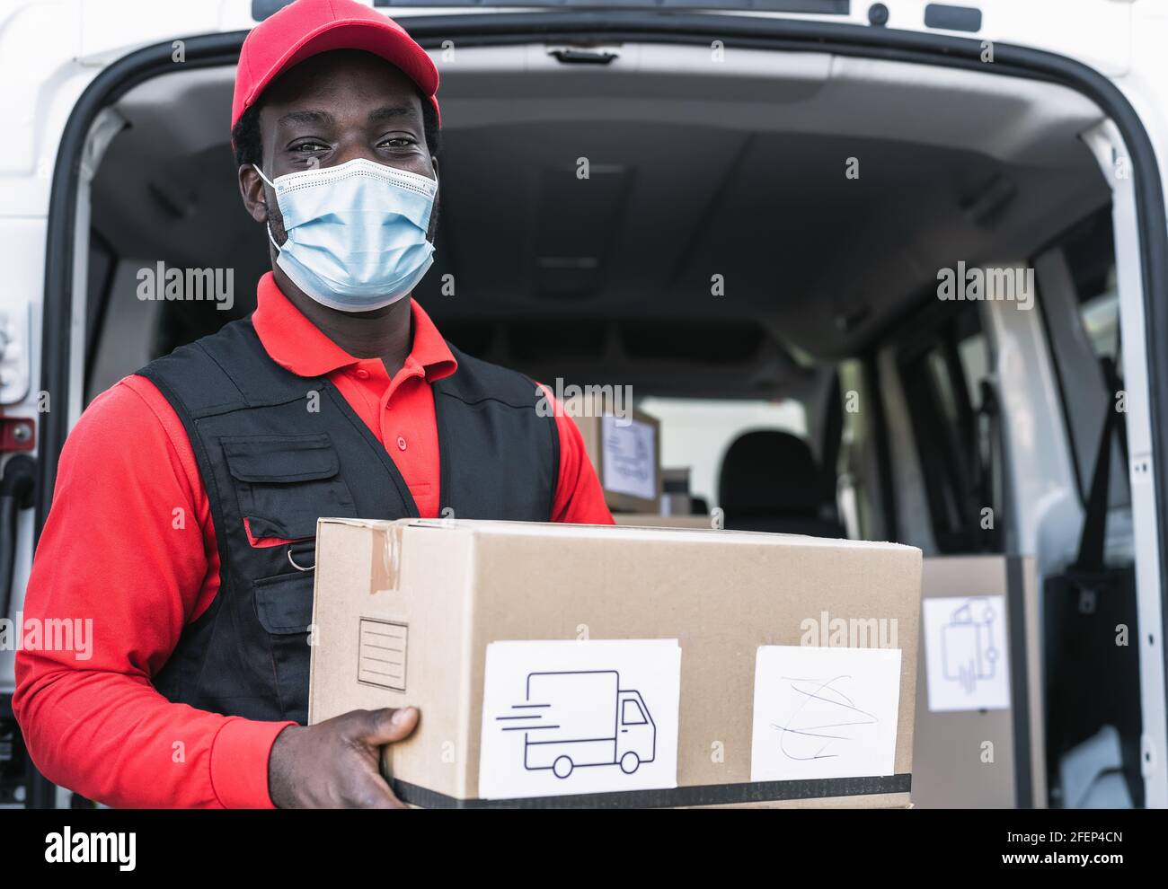 African delivery man carrying cardboard box while wearing face mask to ...