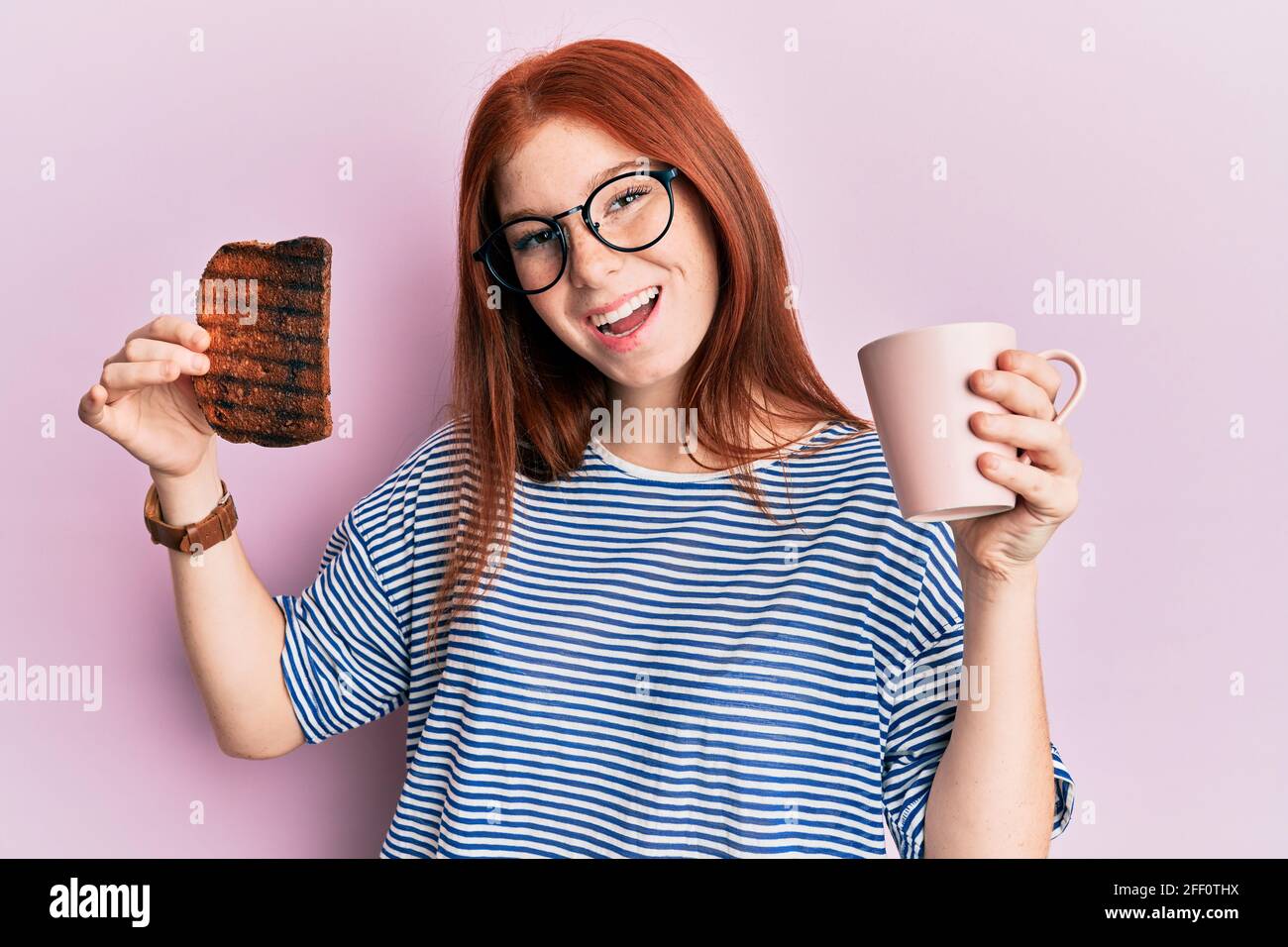 Young red head girl holding burned toast for breakfast smiling and ...