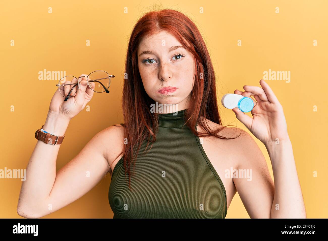 Young red head girl holding glasses and contact lenses puffing cheeks ...