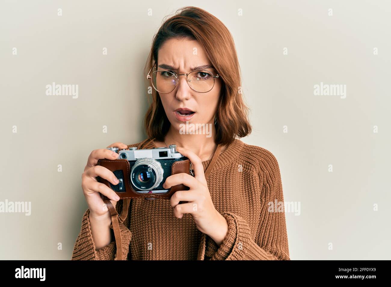 Young caucasian woman holding vintage camera in shock face, looking ...